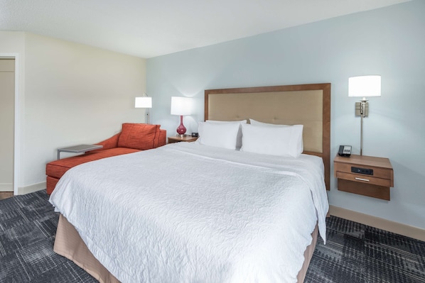 Quarto de hotel com uma cama grande, uma mesa de cabeceira com abajur, um criado-mudo com telefone e uma cadeira vermelha.