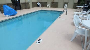 Piscina retangular com marcação de profundidade de 3 pés, rodeada por cadeiras e mesas de plástico branco, e um edifício com fachada de tijolos ao fundo.