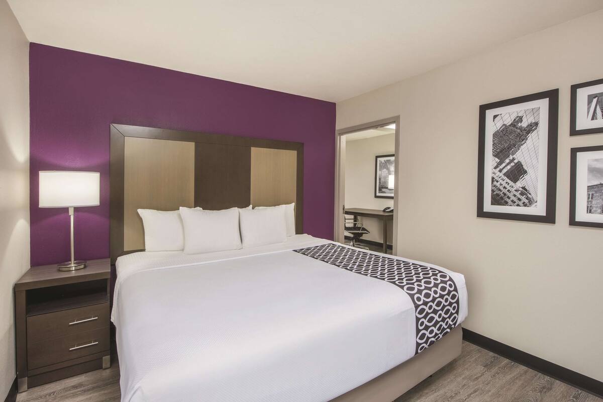 Quarto de hotel com uma cama grande, uma mesa de cabeceira com abajur, cômoda e espelho. Há quadros nas paredes.
