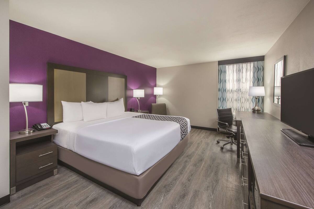 Quarto de hotel com uma cama grande, mesas de cabeceira com luminárias, uma escrivaninha com cadeira e uma televisão.