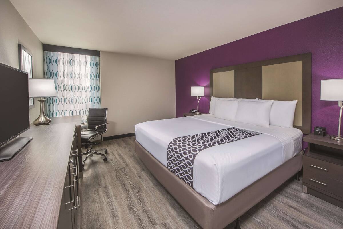 Quarto de hotel com uma cama grande, uma escrivaninha com cadeira e uma janela com cortinas estampadas.
