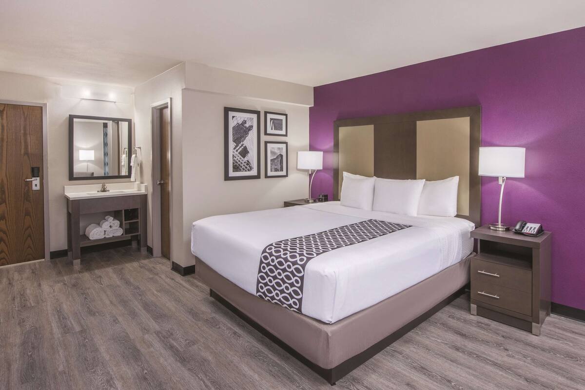 Quarto de hotel com uma cama grande, uma mesa de cabeceira com abajur, uma penteadeira com espelho e uma parede com quadros.