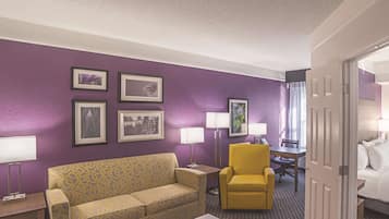 Quarto de hotel com parede roxa, poltrona amarela, sofá, duas mesas de cabeceira com luminárias e uma porta que dá para outro cômodo.