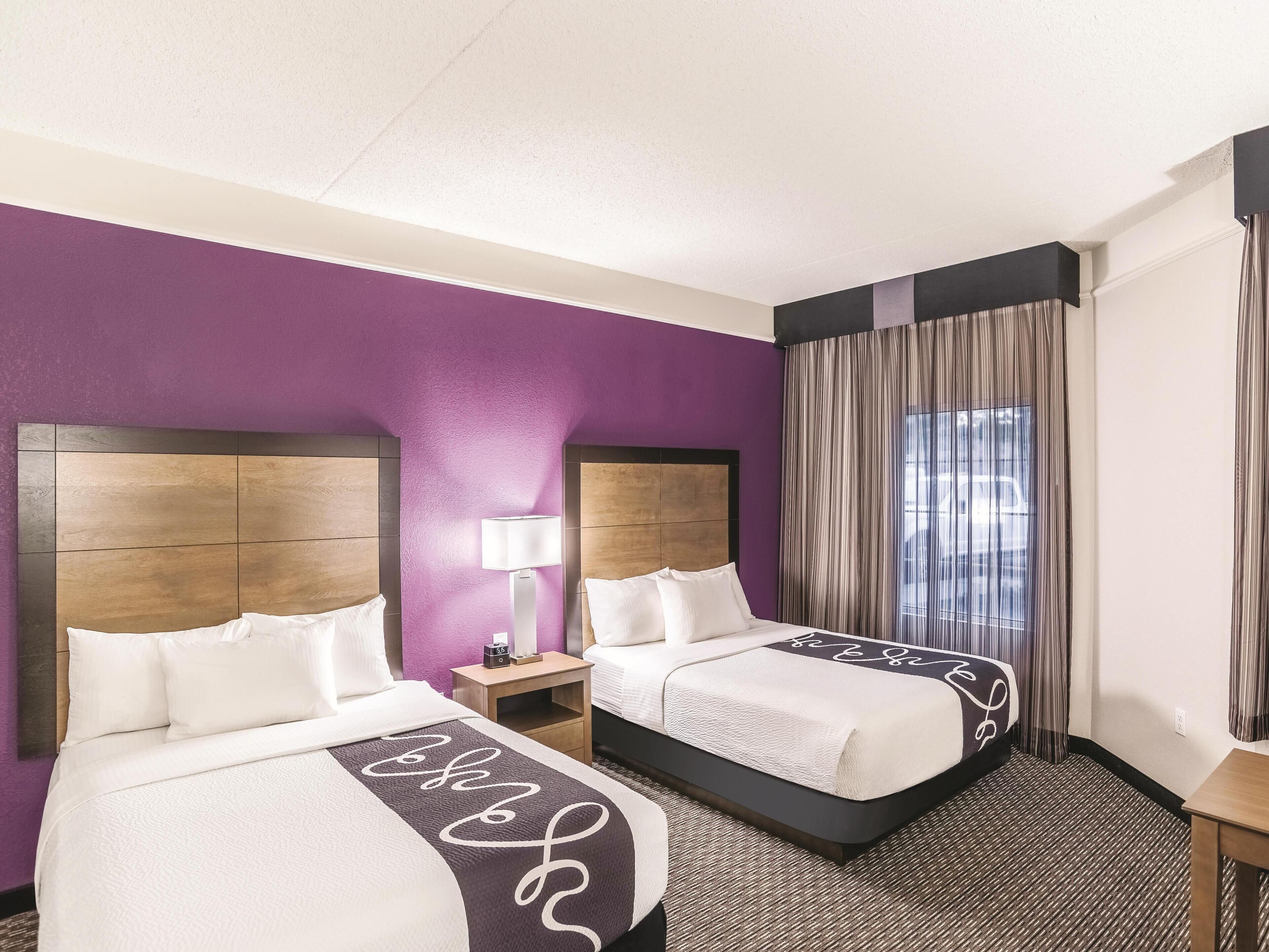 Quarto de hotel com duas camas, uma mesa de cabeceira com abajur e uma janela com cortinas.