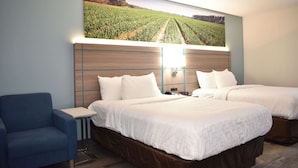 Quarto de hotel com duas camas, uma mesa pequena, uma poltrona azul e uma grande fotografia de um campo na parede.