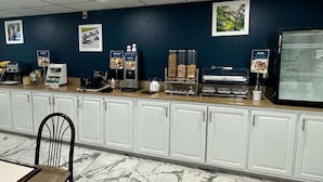 Uma área de pequenos-almoços com uma máquina de café, um balcão refrigerado e uma zona de estar com uma cadeira.