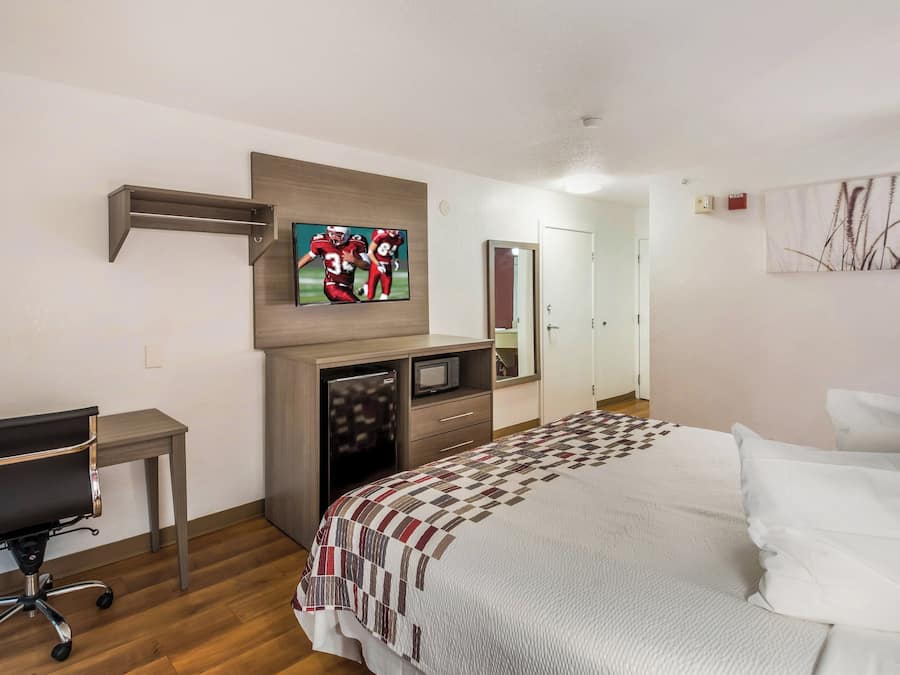 Quarto de hotel com cama, escrivaninha, televisão, refrigerador e porta.