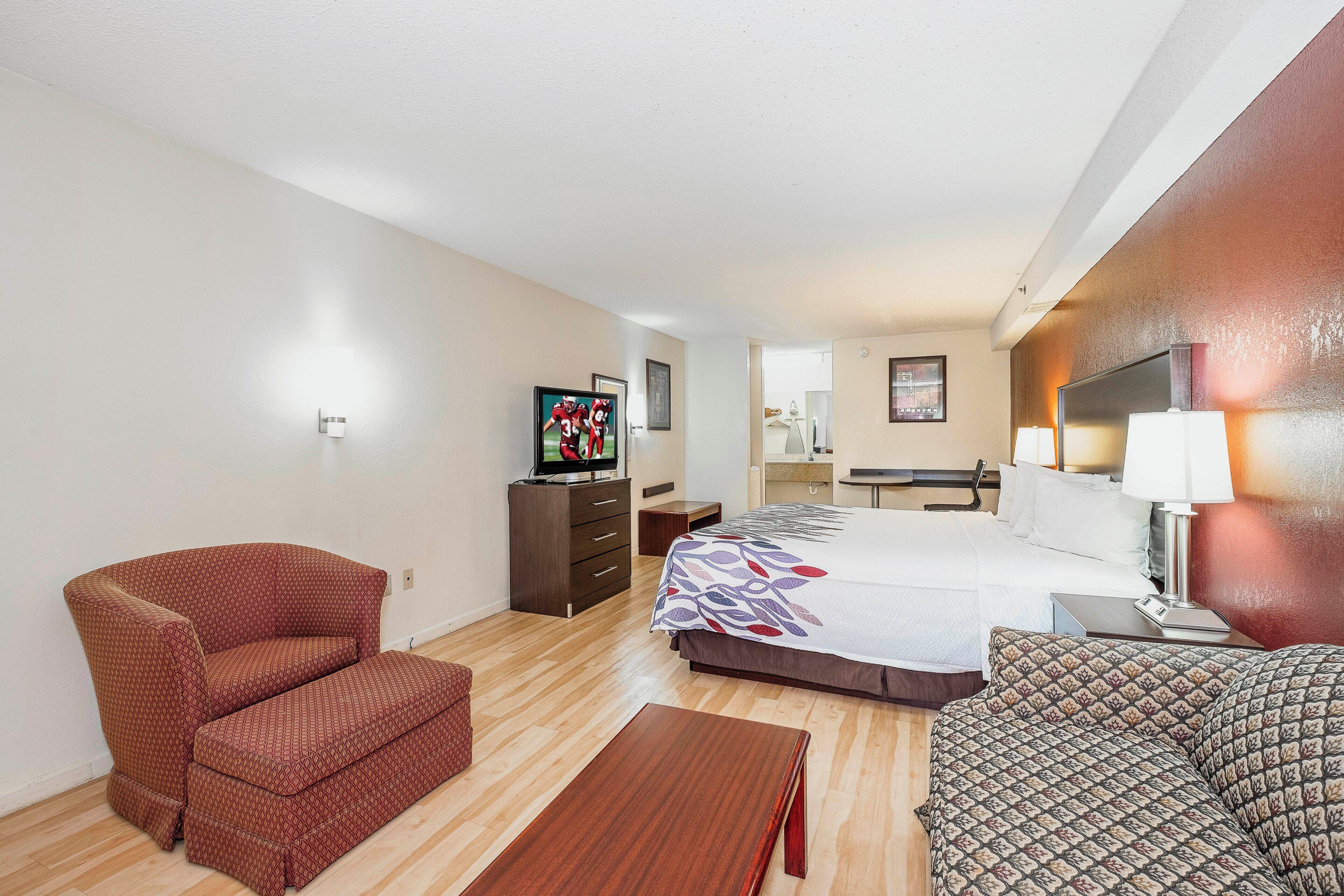 Quarto de hotel com cama, televisão, cadeira, escrivaninha e abajur.