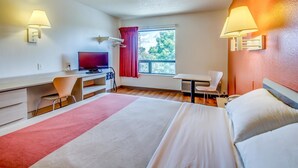 Quarto de hotel com cama, escrivaninha, cadeira, televisão e duas luminárias.