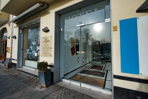 Entrada de hotel com porta de vidro, placa com o nome do hotel e um pequeno vaso de planta.
