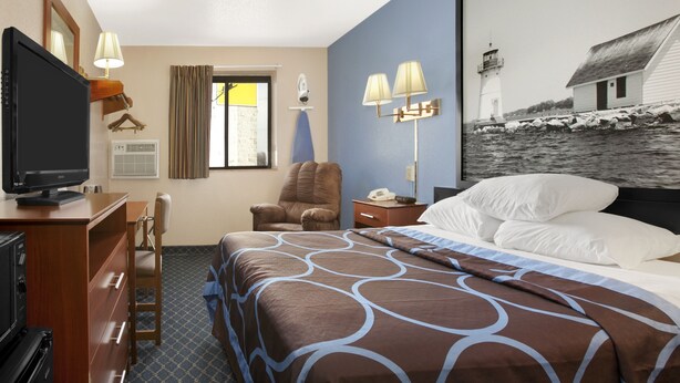 Quarto de hotel com uma cama, uma escrivaninha com televisão, uma cadeira, uma mesinha de cabeceira e uma luminária na parede.