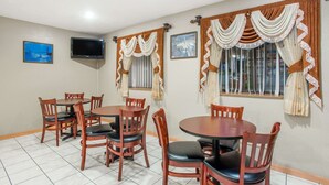 Uma sala de jantar com uma mesa redonda e seis cadeiras de madeira com almofadas pretas. Há duas janelas com cortinas decorativas. Uma televisão de tela plana está fixada na parede acima da mesa.