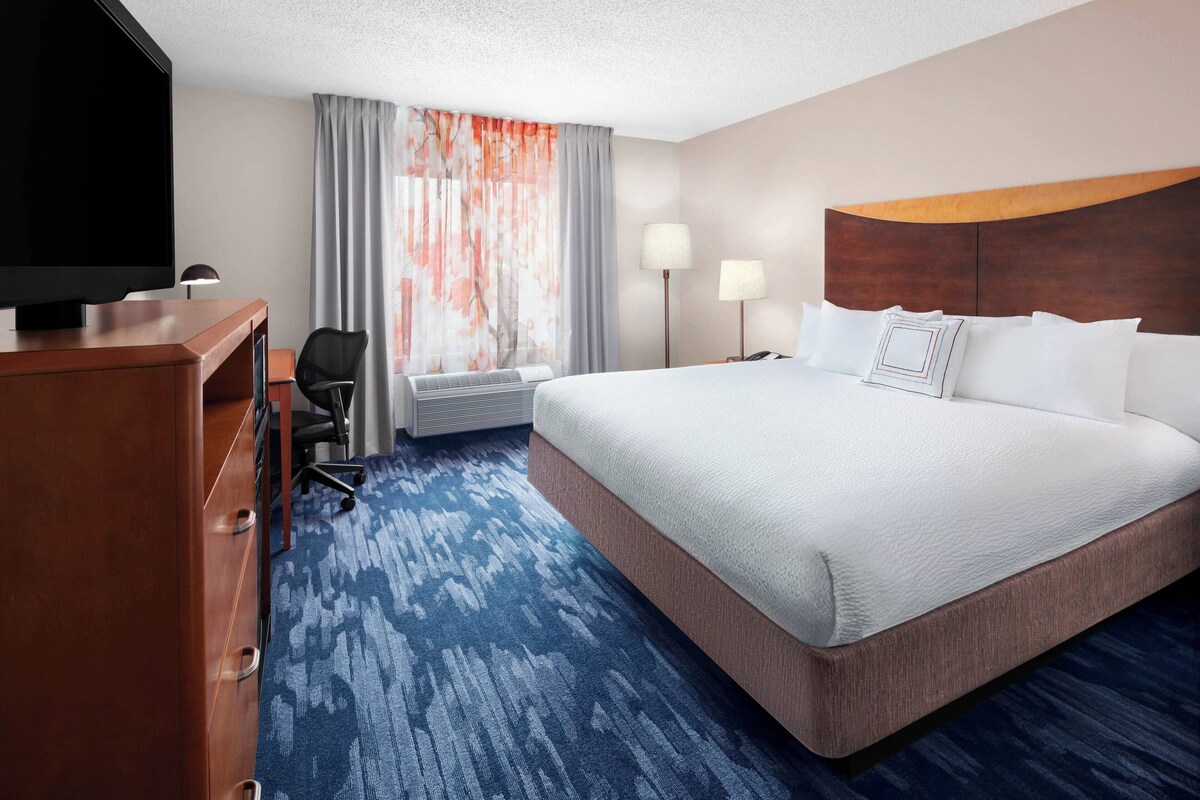 Quarto de hotel com uma cama grande, uma escrivaninha com televisão, uma cadeira e uma janela com cortinas.