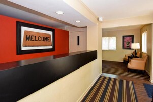 Área de recepção de hotel com um letreiro 'WELCOME', balcão de atendimento, espaço de estar com uma luminária e um carpete listrado.