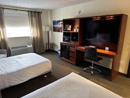 Quarto de hotel com cama, escrivaninha com computador, televisão e janela com cortinas.