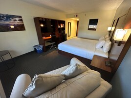 Quarto de hotel com cama, escrivaninha, cadeira, mesa pequena e lixeira.
