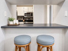 Uma cozinha com bancada escura, eletrodomésticos de aço inoxidável e dois banquetas de madeira com almofadas azuis.
