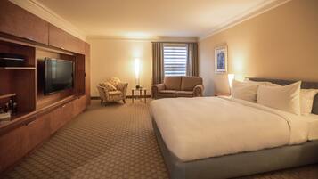 Quarto de hotel com uma cama grande, televisão na parede, uma mesinha, duas poltronas e uma janela com persianas.