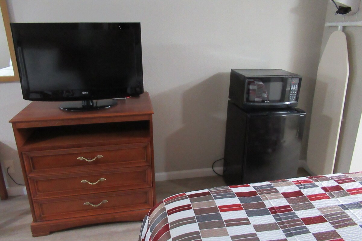 Quarto com cômoda de madeira, televisão de tela plana, micro-ondas preto e cama com colcha xadrez.