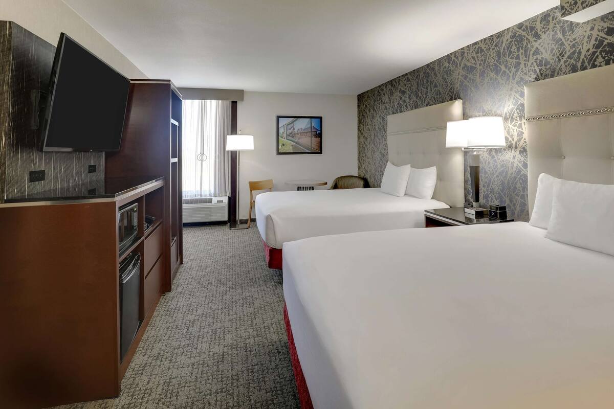 Quarto de hotel com duas camas, uma televisão, uma escrivaninha e um abajur.