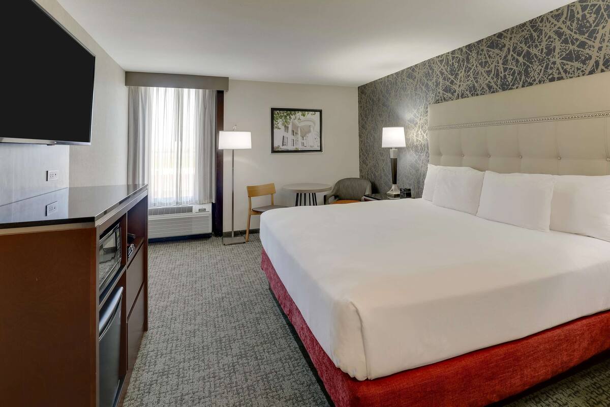 Quarto de hotel com uma cama grande, uma escrivaninha com televisão de tela plana, uma cadeira, uma mesa e uma janela com cortinas.