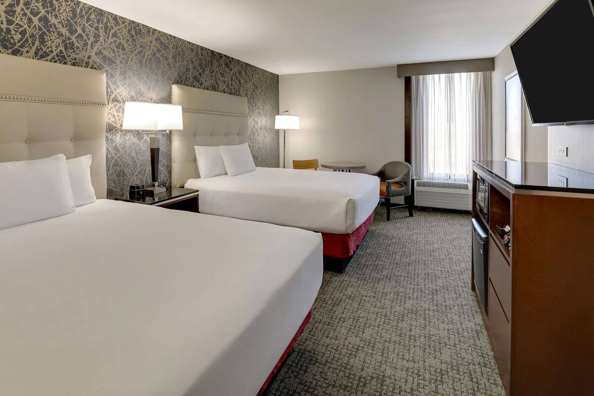 Quarto de hotel com duas camas, uma escrivaninha, uma cadeira, uma televisão e uma janela grande com cortinas.