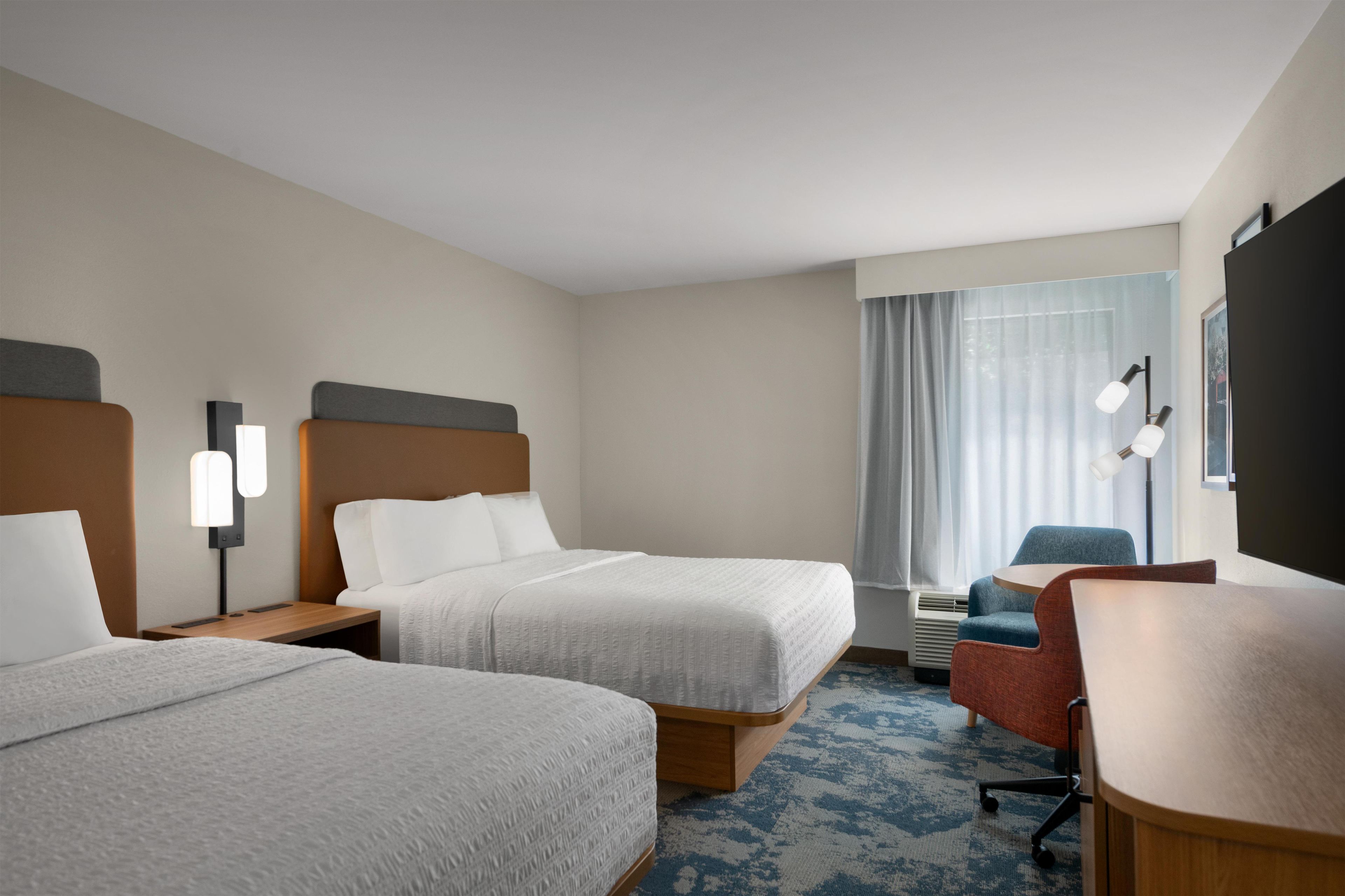 Quarto de hotel com duas camas, uma escrivaninha, uma cadeira e uma janela grande com cortinas.