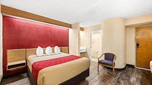 Quarto de hotel com cama, cadeira, cabeceira de madeira, parede com detalhe em vermelho e uma porta.