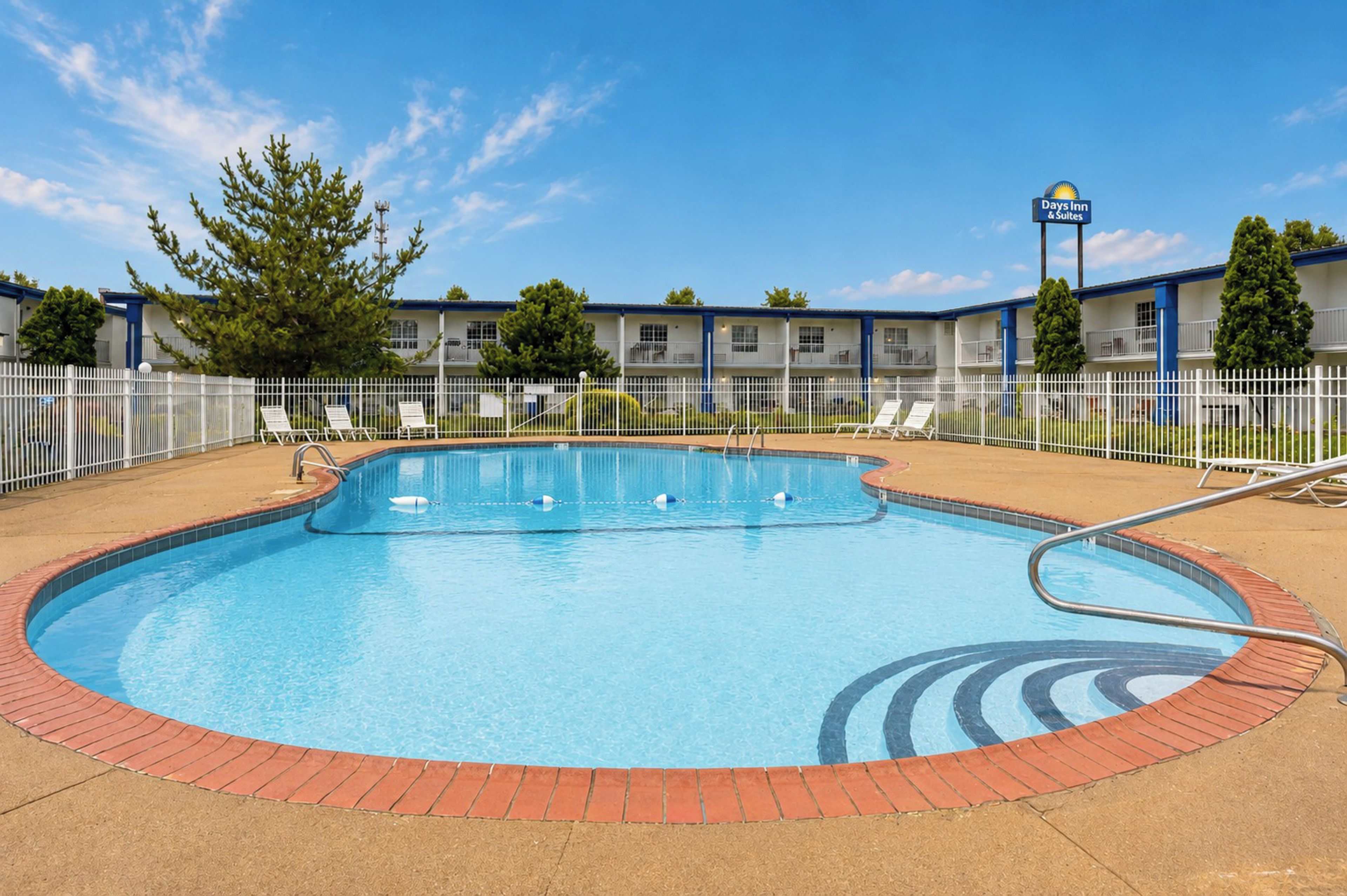 Piscina com borda de tijolos, cercada por uma cerca branca, e um letreiro Days Inn ao fundo.