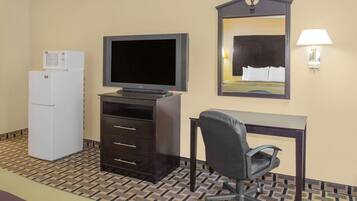 Quarto de hotel com televisão de tela plana, geladeira, escrivaninha com cadeira e espelho na parede.
