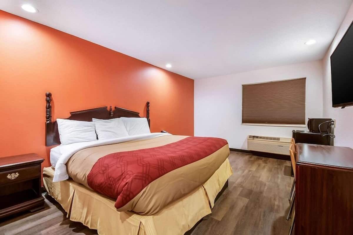 Quarto de hotel com uma cama, mesa de cabeceira, escrivaninha, televisão e janela com persianas.