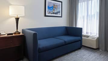 Quarto com sofá azul, escrivaninha com luminária, quadro pendurado na parede e janela com cortinas.