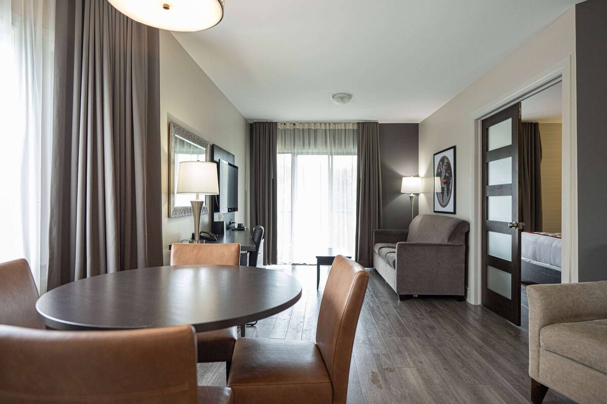 Quarto de hotel com uma mesa de jantar redonda, quatro cadeiras, um sofá, uma televisão e uma porta que dá para outro cômodo.