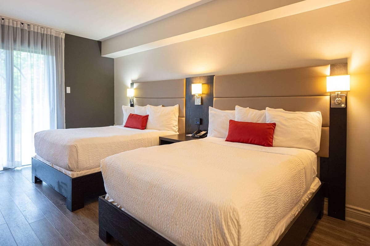 Duas camas de solteiro com roupa de cama branca e almofadas vermelhas, luminárias de cabeceira e piso de madeira.