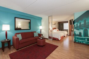 Quarto de hotel moderno com um sofá vermelho, um banquinho vermelho, um espelho grande e uma pintura de um edifício na parede.