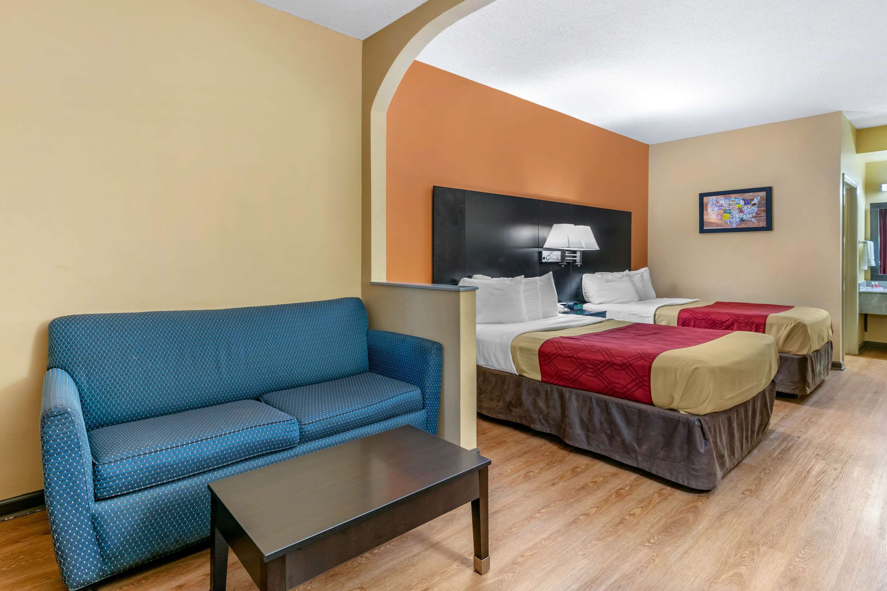 Quarto de hotel com sofá azul, uma pequena mesa, duas camas com roupa de cama vermelha e bege, uma cabeceira e um quadro na parede.