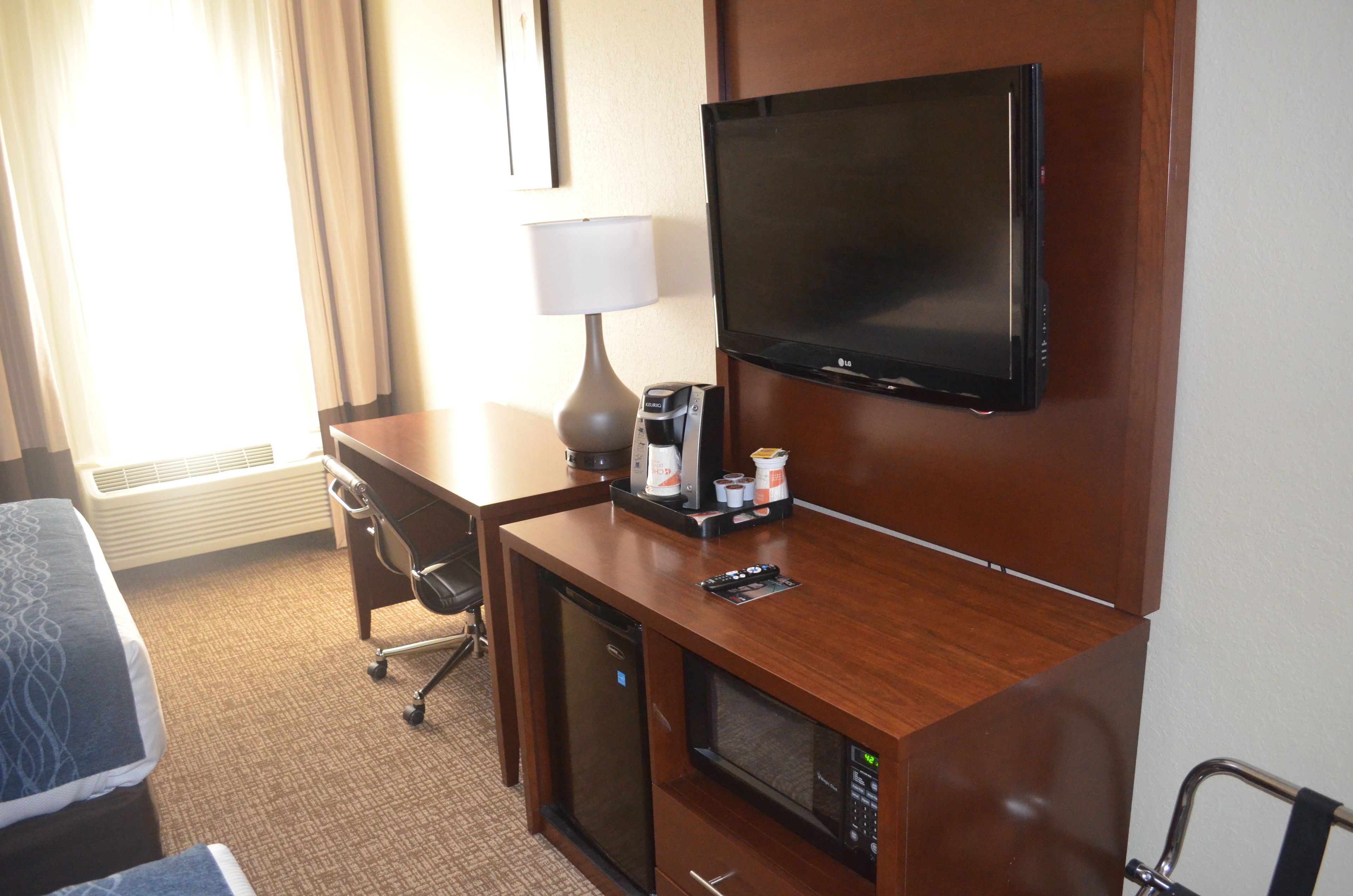 Quarto de hotel com cama, escrivaninha, televisão, luminária, cafeteira e micro-ondas.