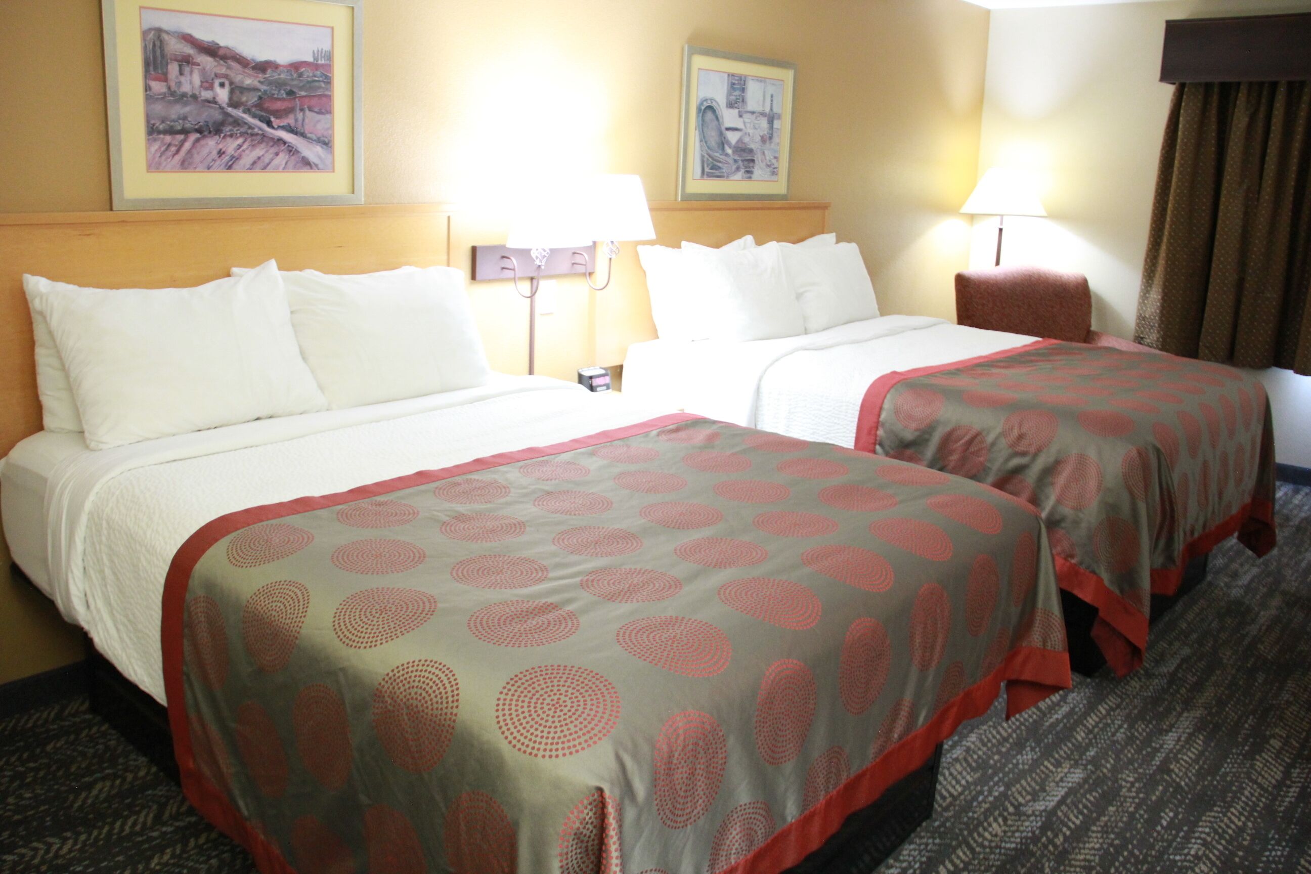 Quarto de hotel com duas camas, cada uma com colcha estampada em vermelho e cinza, almofadas brancas e cabeceira. Há duas luminárias fixadas na parede, um quadro emoldurado e uma janela com cortinas.