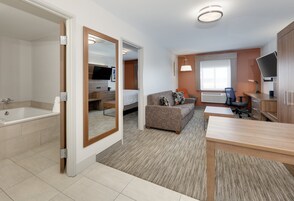 Quarto de hotel moderno com banheiro, área de estar com sofá e TV, e uma escrivaninha com cadeira.