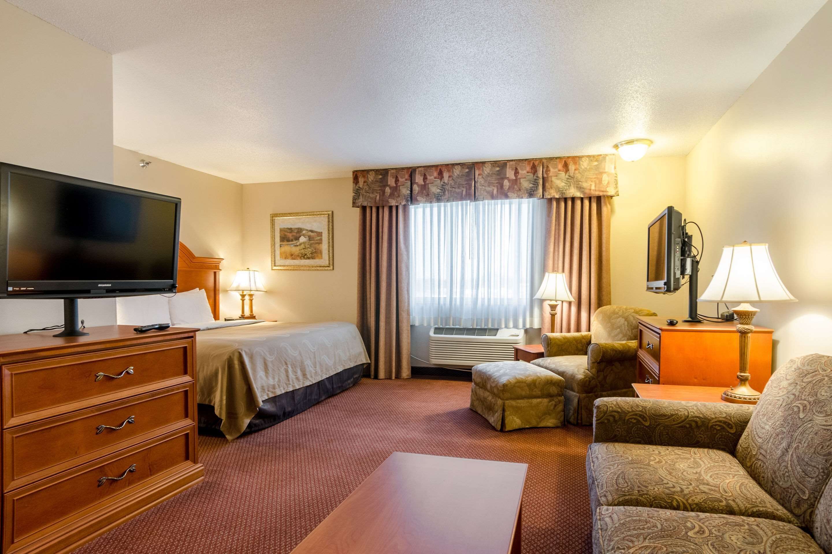 Quarto de hotel com cama, televisão, escrivaninha, poltrona, sofá, cômoda e janela com cortinas.