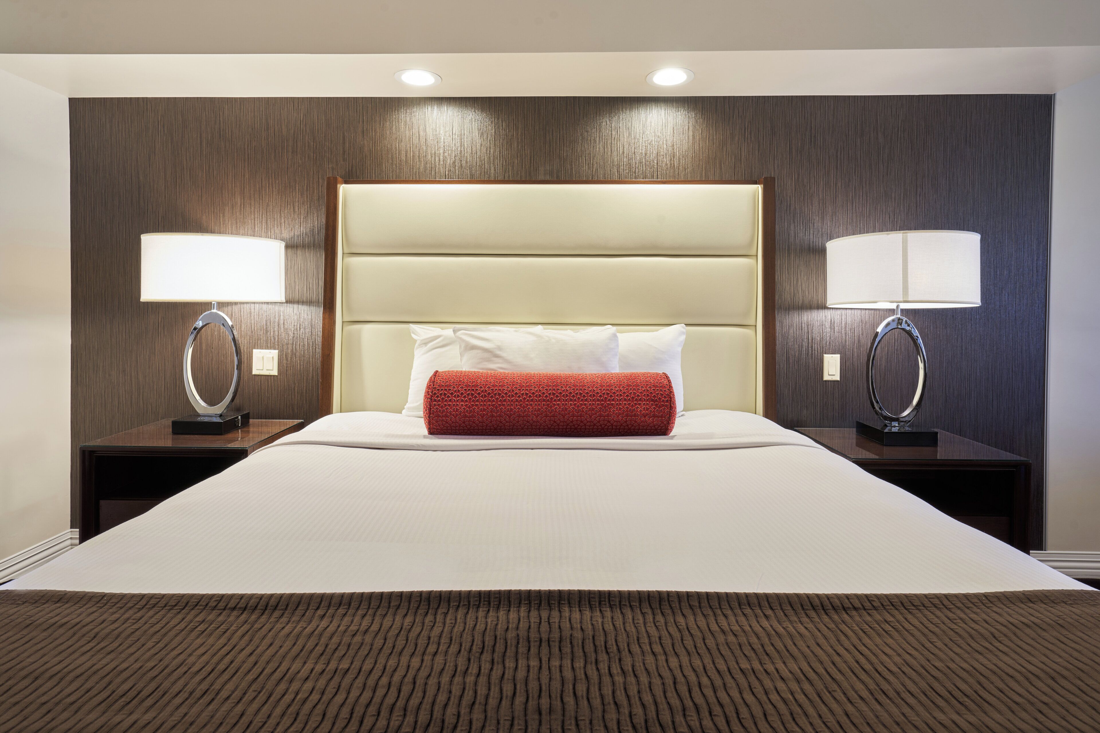Quarto de hotel com uma cama grande, duas mesas de cabeceira com abajures, uma cabeceira e um tapete marrom.