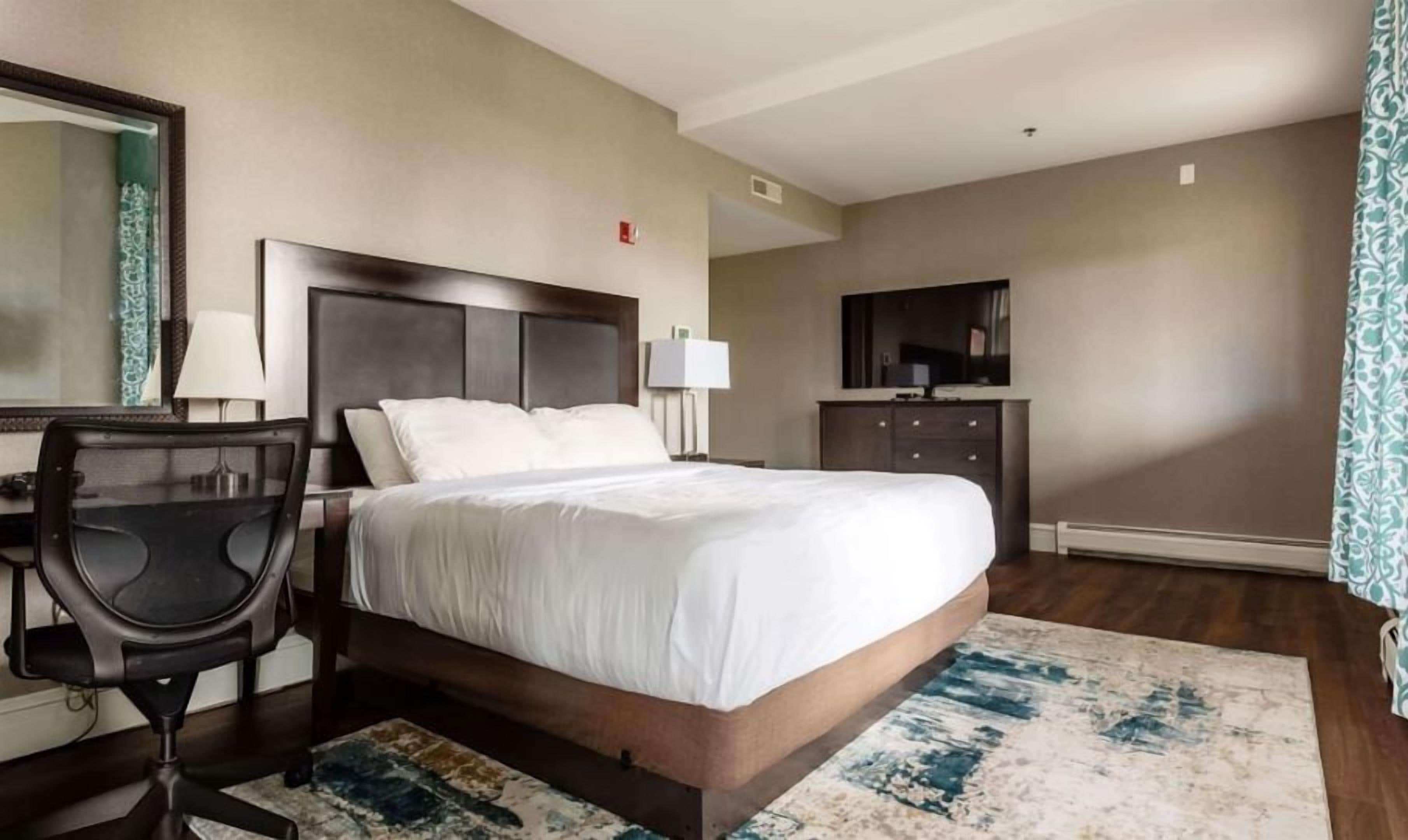 Quarto de hotel com cama, escrivaninha, cadeira, televisão e espelho.