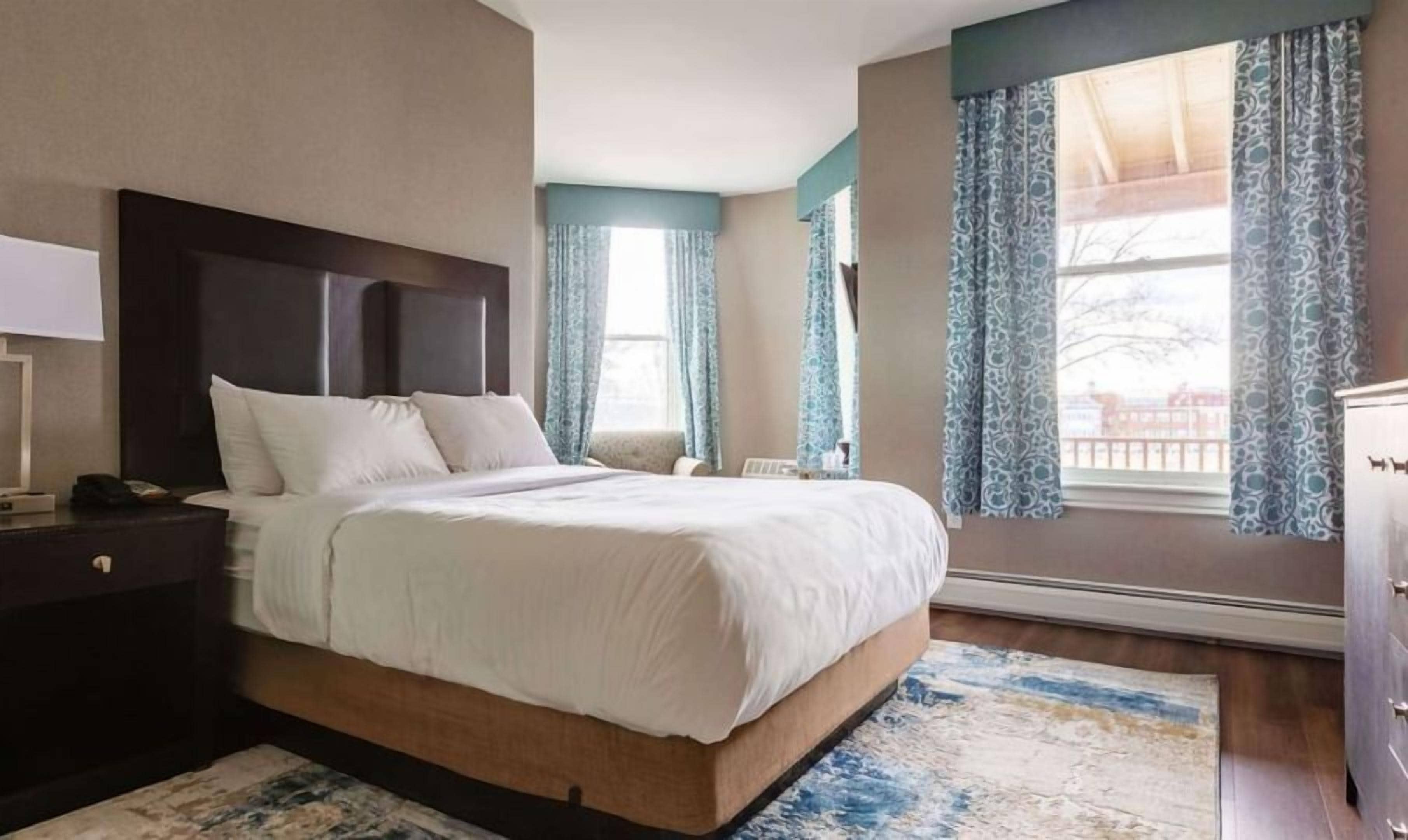 Quarto de hotel com uma cama grande, mesa de cabeceira, cômoda e janela com cortinas.