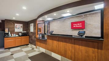 Uma área de recepção de hotel com balcão de check-in, máquina de café e placa indicando Wi-Fi gratuito em todos os quartos.