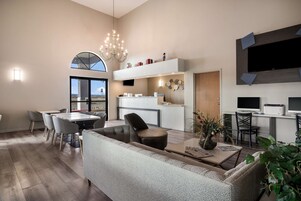 Uma sala moderna com sofá, mesa de centro e área de jantar. Há uma cozinha com bancada e uma televisão grande fixada na parede.
