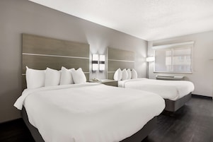 Quarto de hotel com duas camas, um espelho grande, uma mesa de cabeceira com abajur e uma janela com persianas.