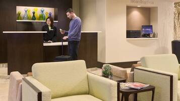Uma área de recepção de hotel com um balcão, um homem e uma mulher atrás dele, uma área de estar com poltronas, uma pequena mesa com uma revista e uma placa na parede com a inscrição 'Boarding Pass'.