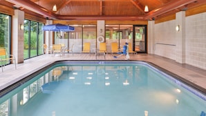 Piscina interna com paredes azulejadas, vigas de madeira no teto e vários cadeiras de descanso e guarda-sóis.