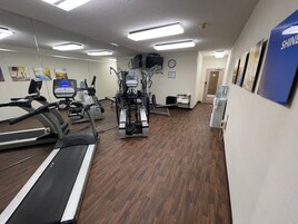 Uma academia com esteiras, elípticos e uma bicicleta ergométrica. Há relógios fixados na parede e um bebedouro.