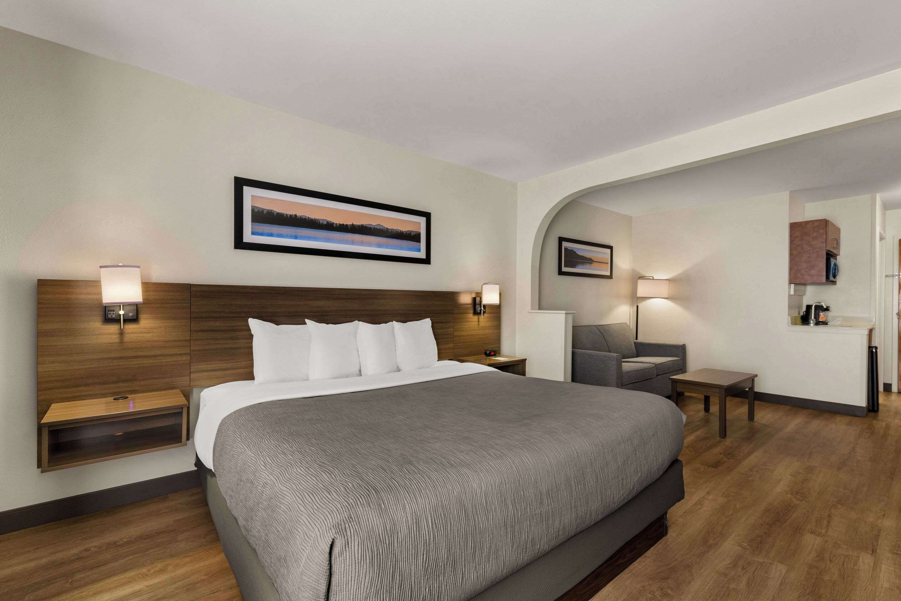 Quarto de hotel com uma cama grande, uma escrivaninha, uma cadeira e uma mesinha. Há duas luminárias na parede e um quadro.